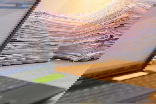 Computer notebook and documents on desk stack up high waiting to be managed