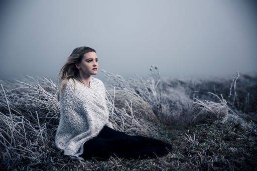 A cute blond girl wearing a knitted scarf sitting in a field covered in ...
