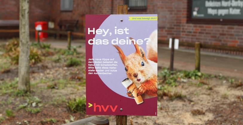 In a flower bed behind a bus stop there is a sign from the Hamburger ...